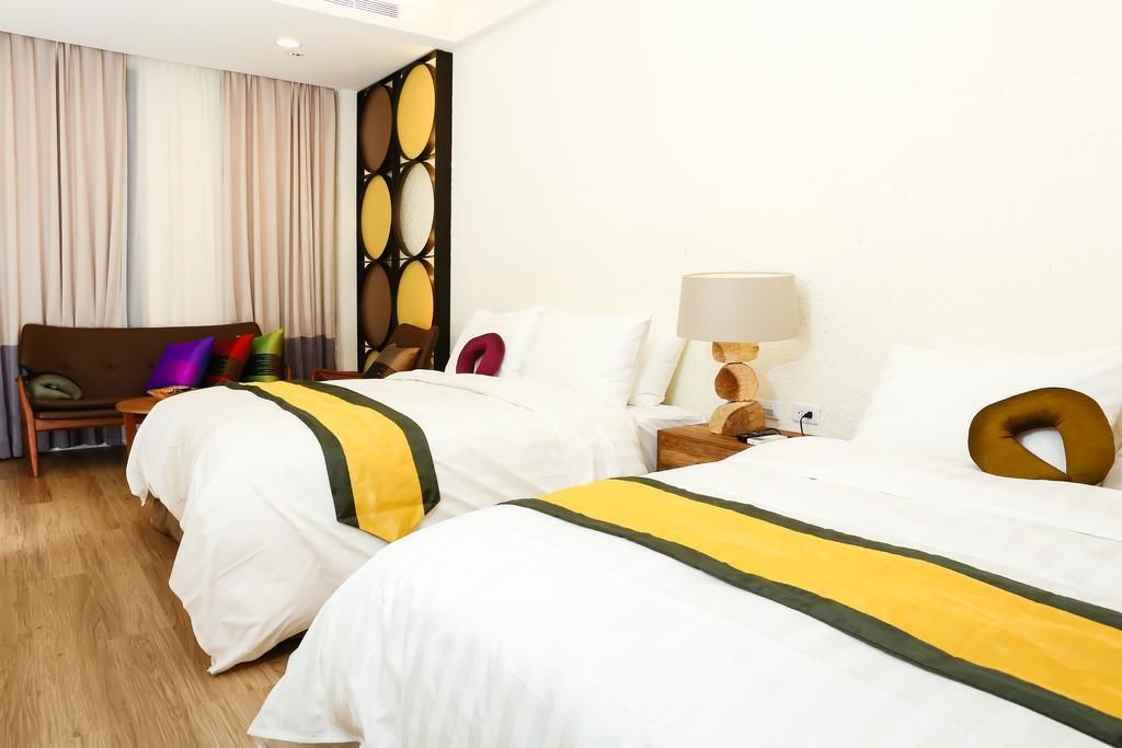 Quarto de hotel com duas camas, uma mesa de cabeceira com abajur, uma cadeira e um painel decorativo na parede.