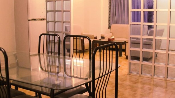 Uma sala de jantar com uma mesa de vidro e cadeiras pretas, um frigorífico e um sofá ao fundo.