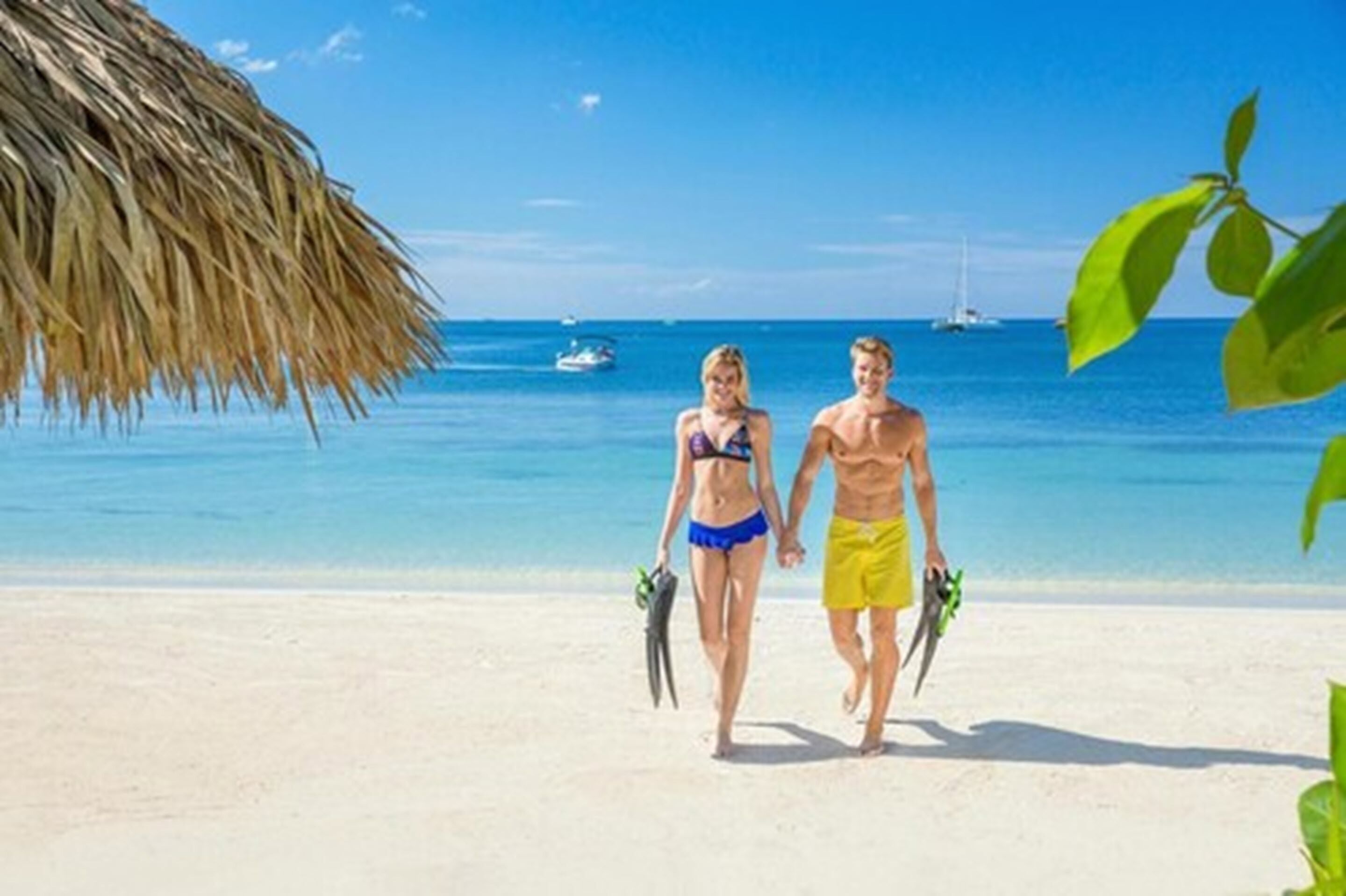 Um casal caminhando na praia, com vista clara do oceano e barcos ao longe.