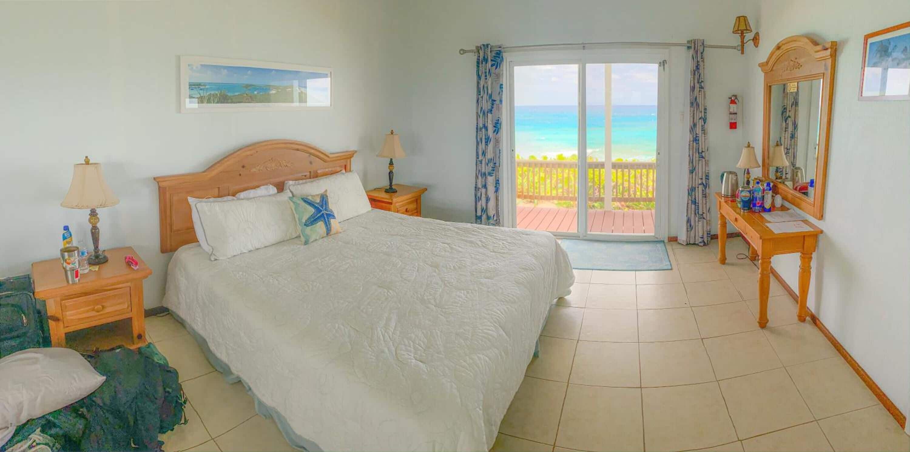 Quarto com cama, mesas de cabeceira, penteadeira, espelho e varanda com vista para o mar.