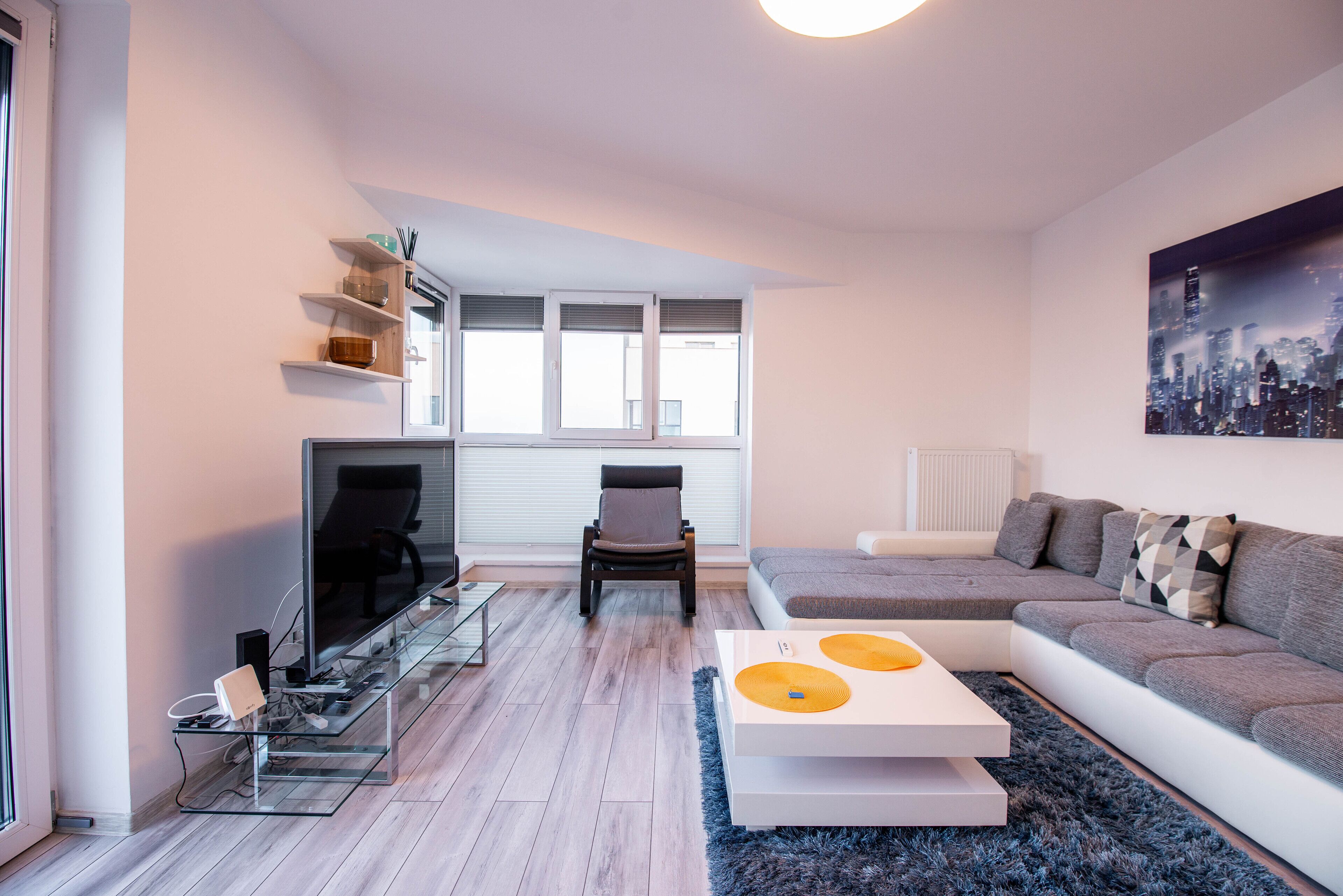 Uma sala de estar moderna com uma mesa de centro de vidro, um sofá cinzento, um sofá branco, uma poltrona preta e uma pintura com vista para a cidade na parede.