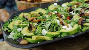 Uma salada de folhas verdes variadas com fatias de abacate, queijo feta e carne grelhada.