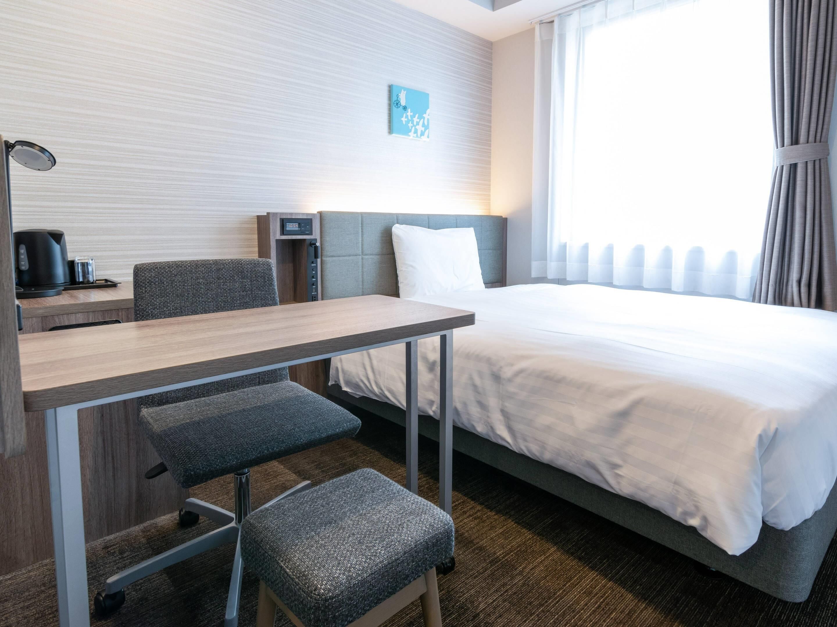 Quarto de hotel com cama, escrivaninha, cadeira, cafeteira e relógio de parede.