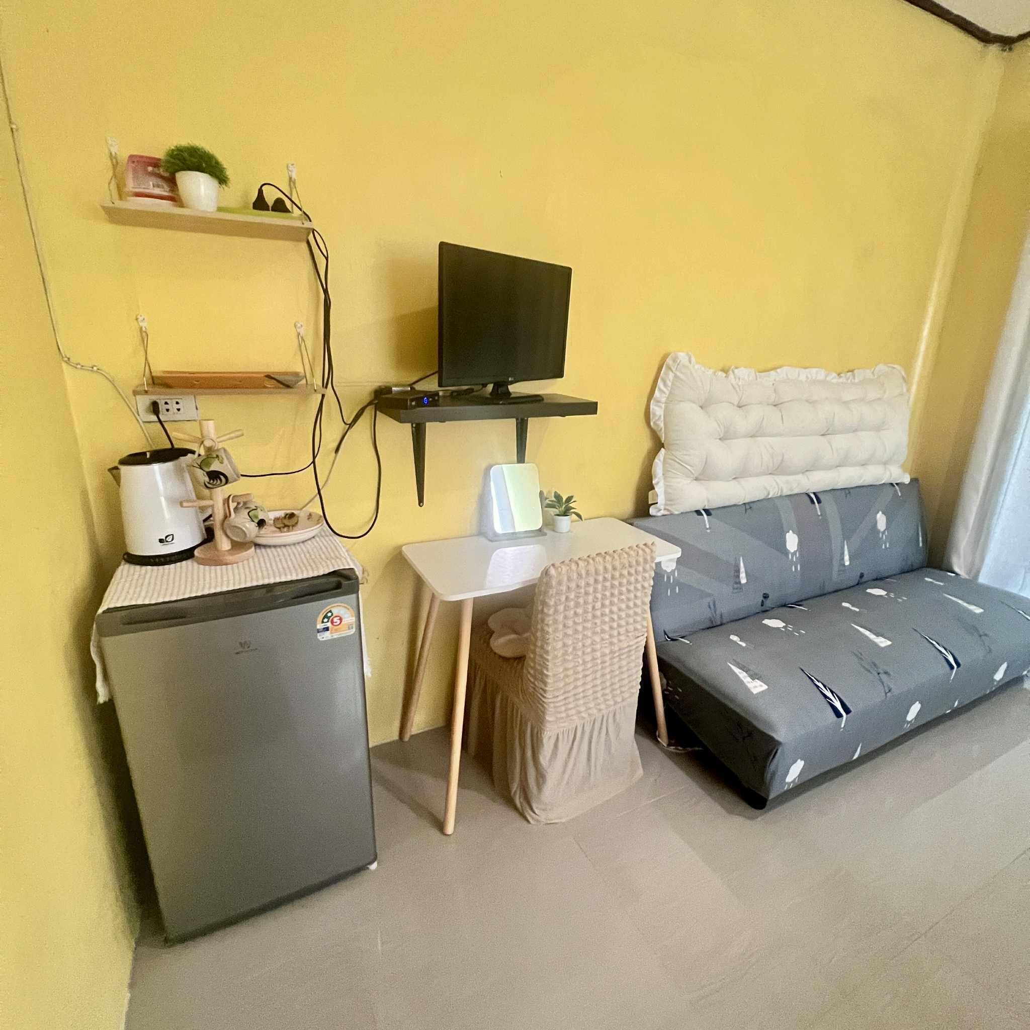 Quarto compacto com cama, escrivaninha pequena, geladeira e televisão.