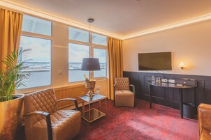 Quarto de hotel com uma janela grande, televisão, mesa com abajur, cadeira e tapete vermelho.