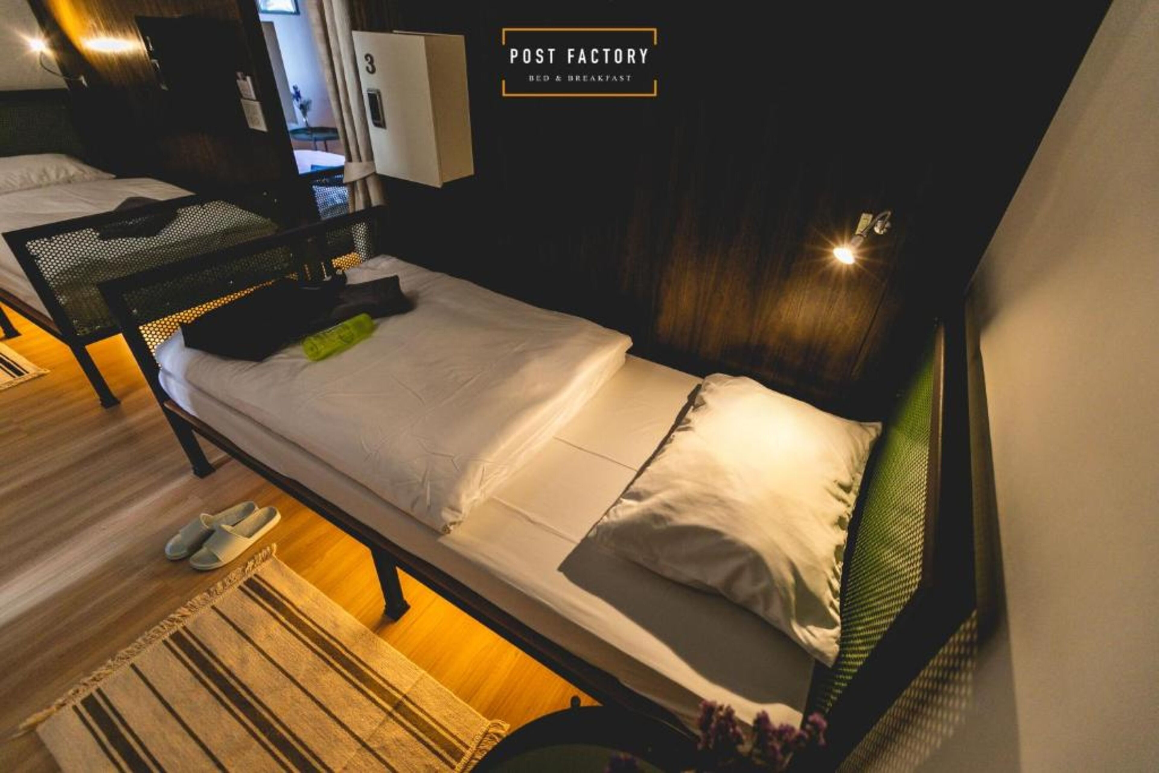 Cama de solteiro em quarto com parede escura e janela pequena. Há um travesseiro verde na cama, e um par de chinelos no chão. O quarto possui piso de madeira e um tapete pequeno.