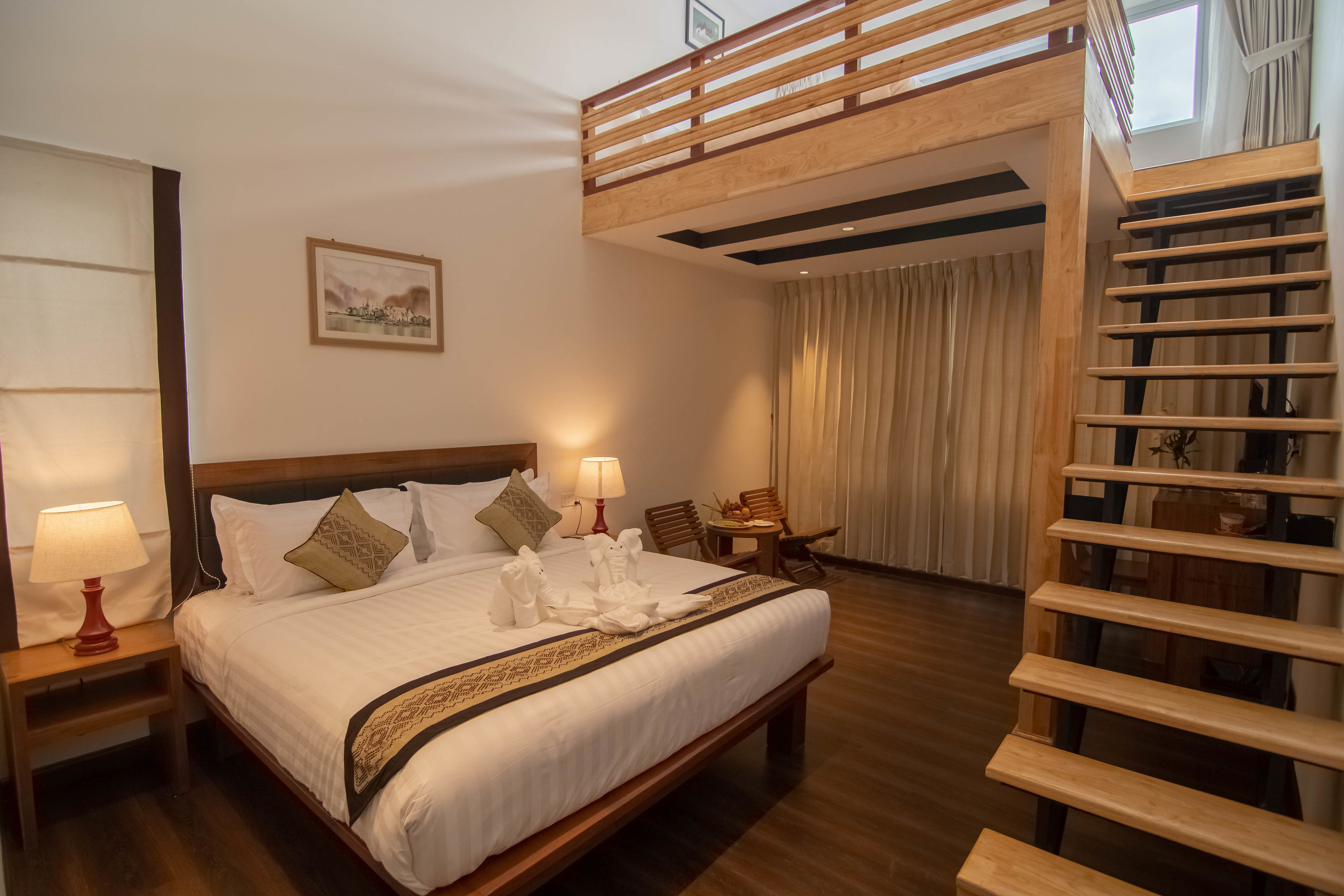 Quarto de hotel com uma cama grande, cabeceira de madeira, mesa de cabeceira com abajur, pequena mesa com frutas e escada de madeira.