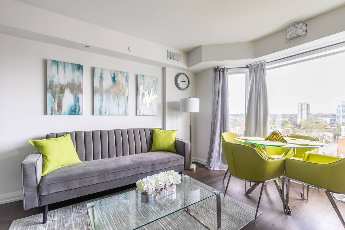 Uma sala de estar moderna com um sofá cinzento, almofadas verdes, uma mesa de centro de vidro e vista para a cidade através de grandes janelas.