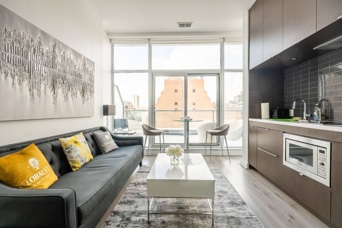 Uma sala de estar moderna com um sofá cinzento, uma mesa de centro branca e uma janela grande com vista para a cidade.