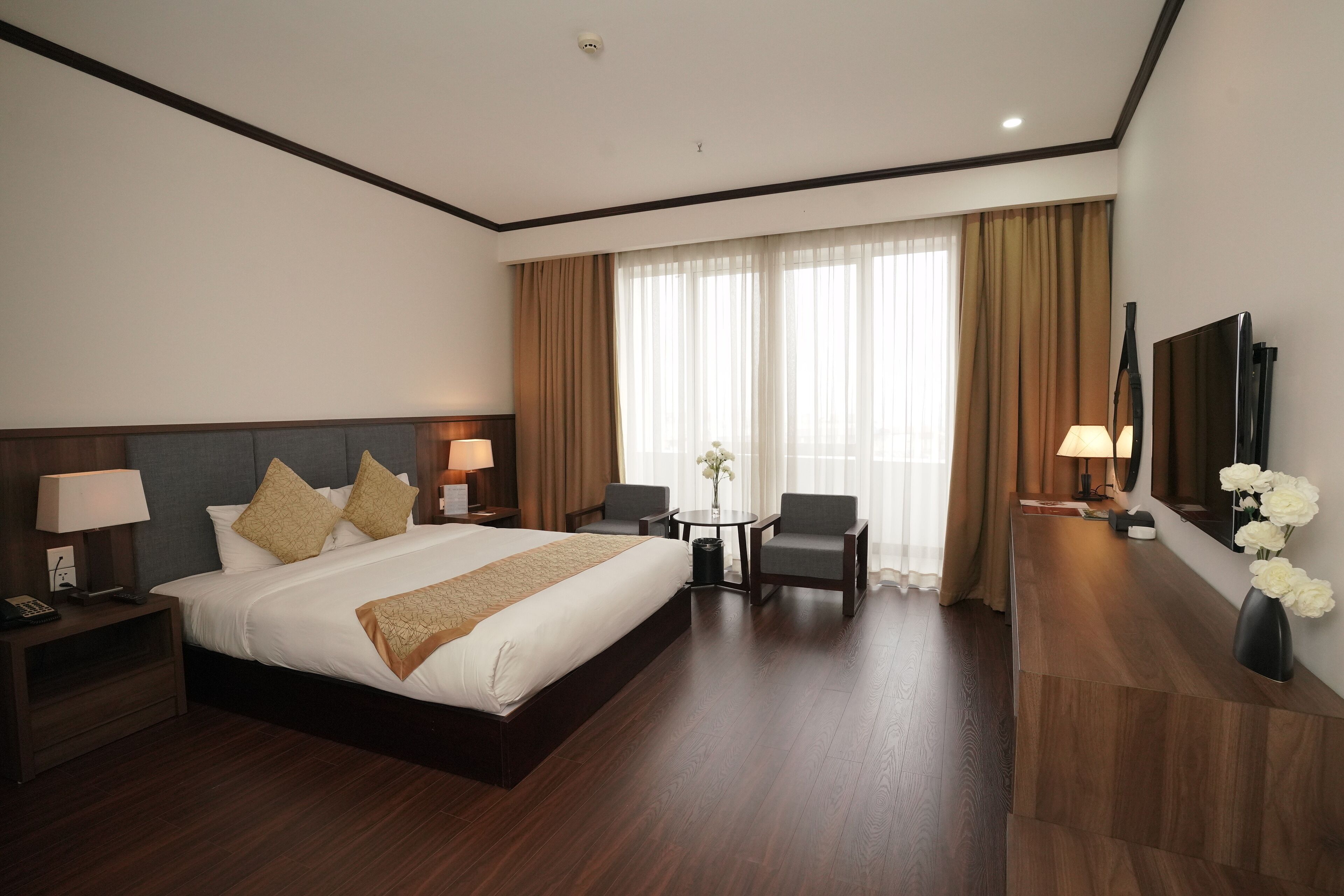 Quarto de hotel com uma cama grande, duas mesas de cabeceira com abajures, uma escrivaninha com cadeira, televisão e uma janela grande com cortinas.