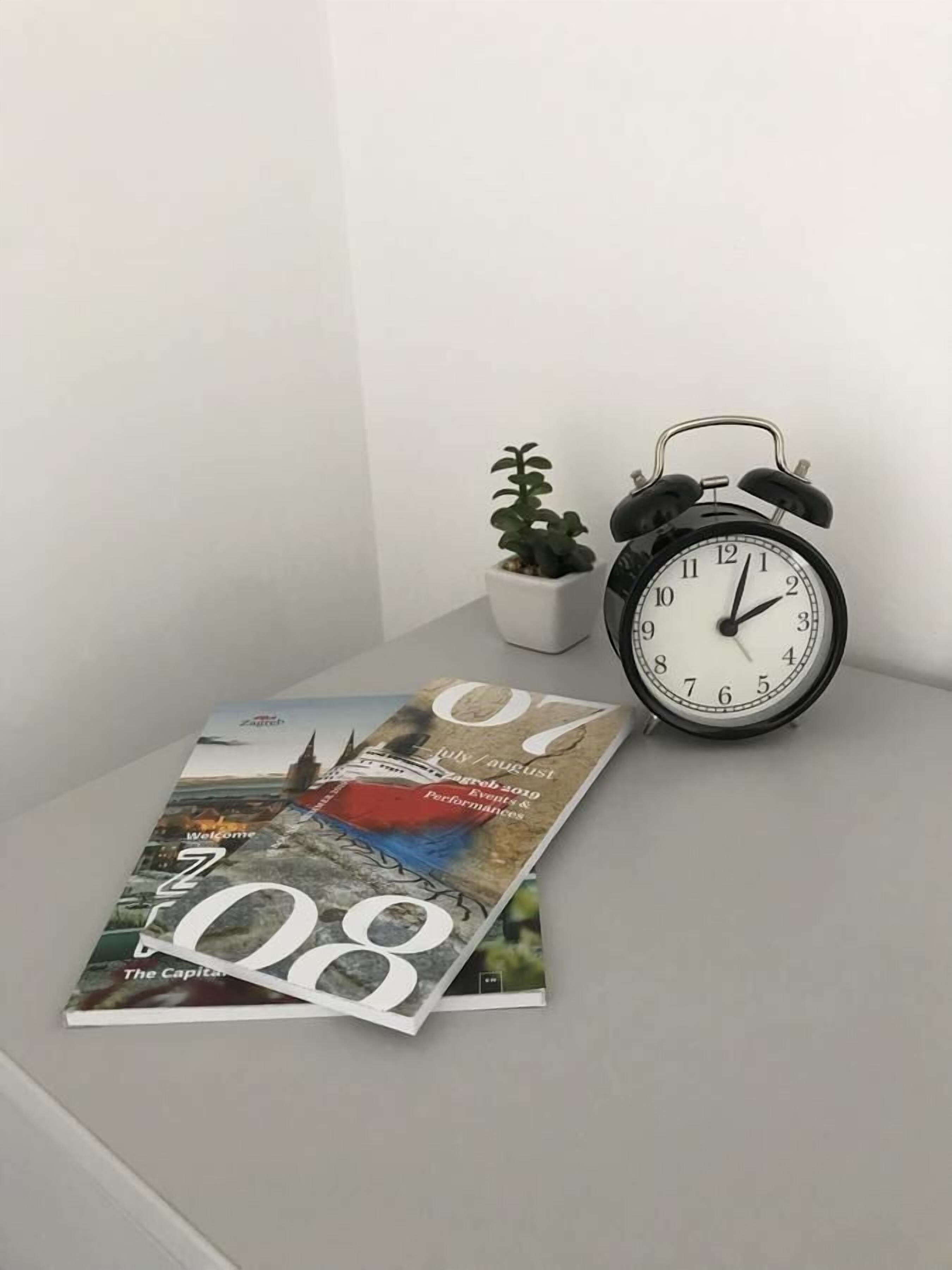 Um despertador preto sobre uma superfície branca, ao lado de uma pequena planta em vaso e uma revista com a capa mostrando uma paisagem urbana.
