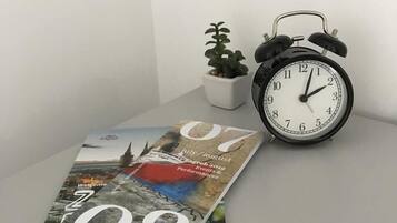 Um despertador preto sobre uma superfície branca, ao lado de uma pequena planta em vaso e uma revista com a capa mostrando uma paisagem urbana.