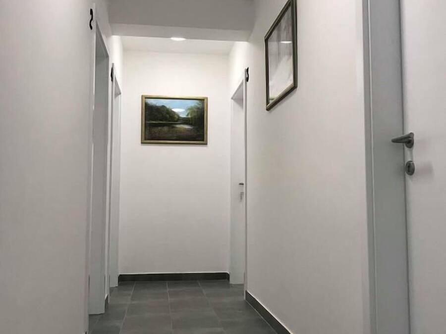 Corredor estreito com paredes brancas e chão em piso escuro. Há duas portas à direita, uma delas aberta. Um quadro emoldurado está pendurado na parede acima da porta aberta.