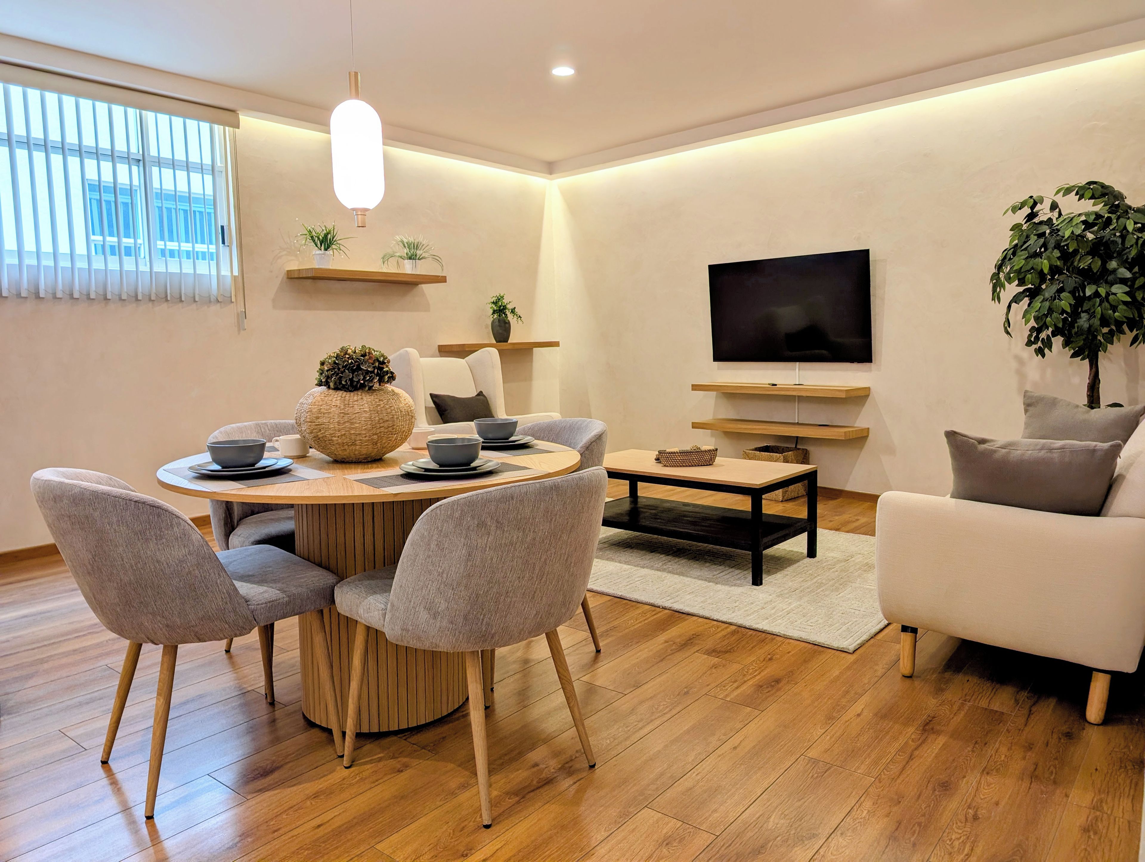 Uma sala de estar moderna com uma mesa de jantar redonda, cadeiras estofadas cinzentas, uma TV de tela plana na parede e um vaso de planta.