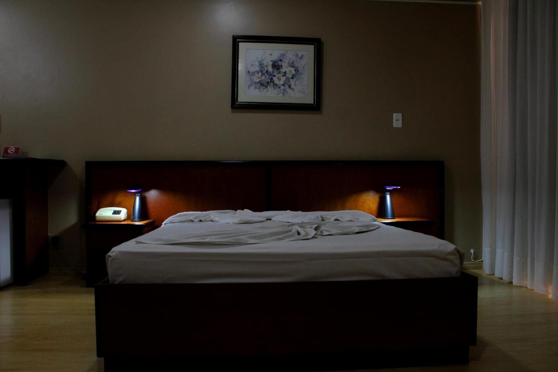 Quarto de hotel com uma cama grande, duas mesas de cabeceira com luminárias, uma obra de arte emoldurada na parede e uma cortina na janela.