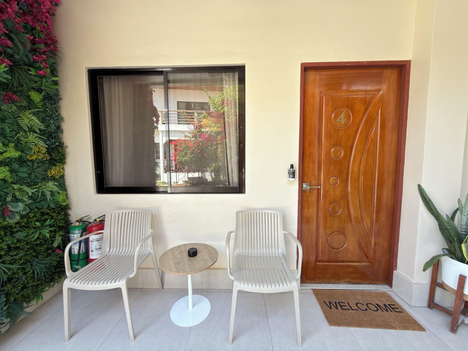 Quarto com duas cadeiras brancas, uma mesa redonda pequena, uma porta de madeira com número e uma janela com vista para edifícios e árvores.