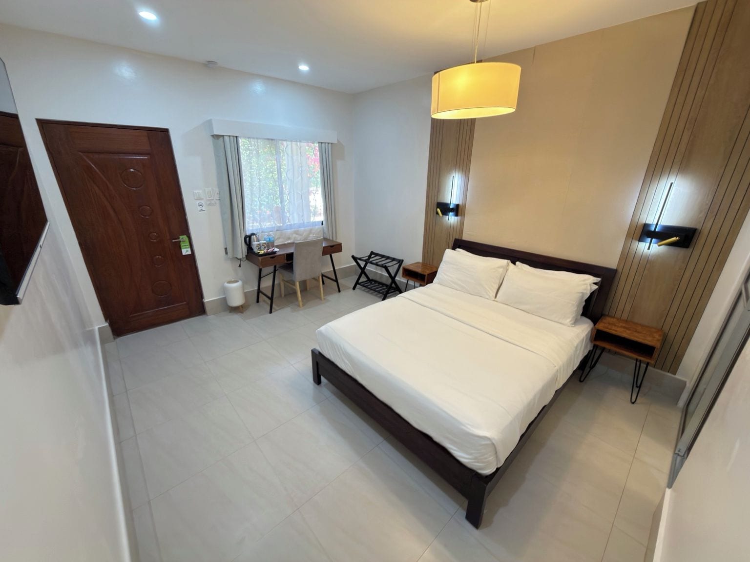 Quarto de hotel com uma cama grande, uma mesa com cadeira, janela com cortinas e luminária suspensa.