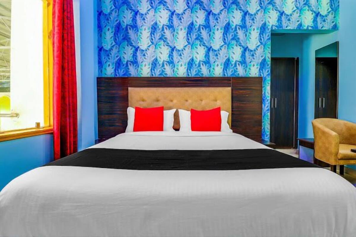 Quarto de hotel com uma cama grande, almofadas vermelhas e brancas, cabeceira de madeira e parede com padrão azul.
