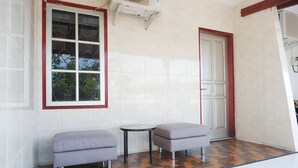Uma área de espera com dois bancos acolchoados cinzentos, uma pequena mesa redonda e uma porta com moldura vermelha.