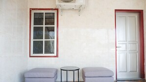 Quarto com duas áreas de estar acolchoadas em cinza, uma pequena mesa redonda, uma janela com moldura vermelha e uma porta branca com moldura vermelha.