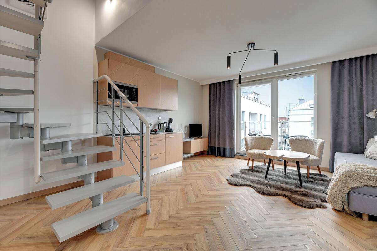 Apartamento moderno com piso em padrão herringbone de madeira, escada, cozinha com armários de madeira, área de jantar com duas cadeiras, sofá e janela grande com cortinas.