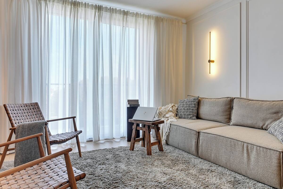 Uma sala de estar moderna com sofá, duas poltronas, um laptop sobre uma mesinha e uma janela grande com cortinas leves.