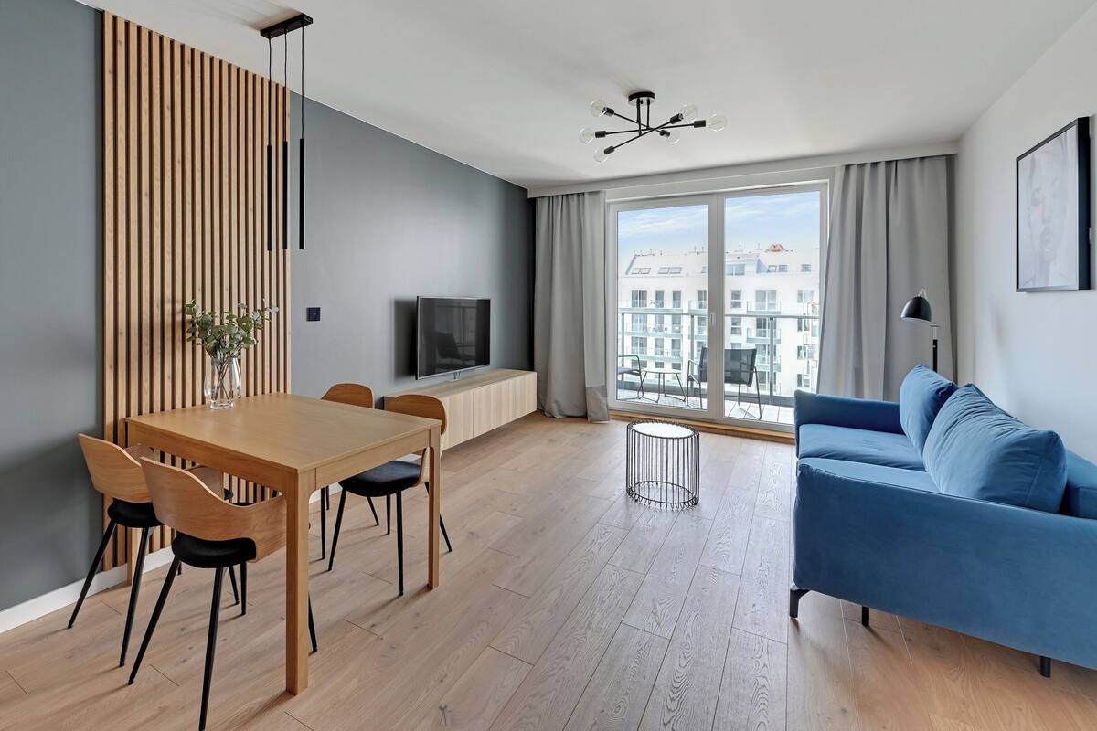 Uma sala de estar moderna com uma mesa de jantar de madeira, sofá azul e uma TV sobre uma prateleira baixa.