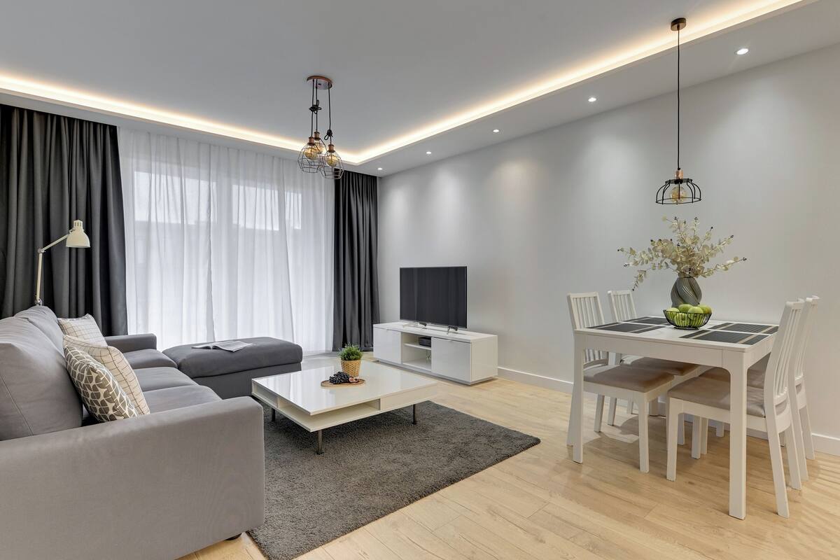 Uma sala de estar moderna com um sofá cinzento, uma mesa de centro branca, uma mesa de jantar com cadeiras, uma televisão e um candeeiro.