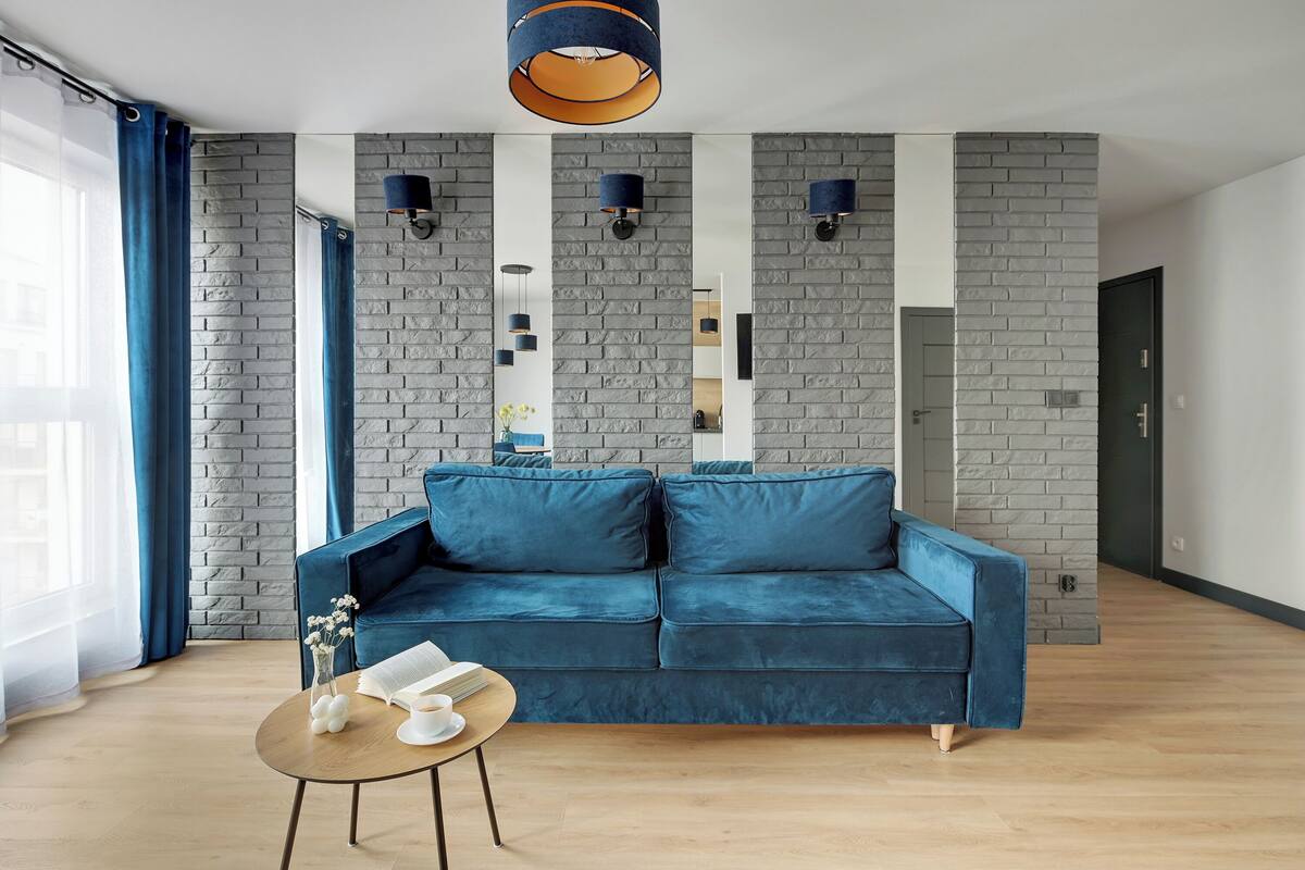 Uma sala de estar moderna com um sofá azul, uma mesa de centro redonda de madeira e uma parede com padrão de tijolos.