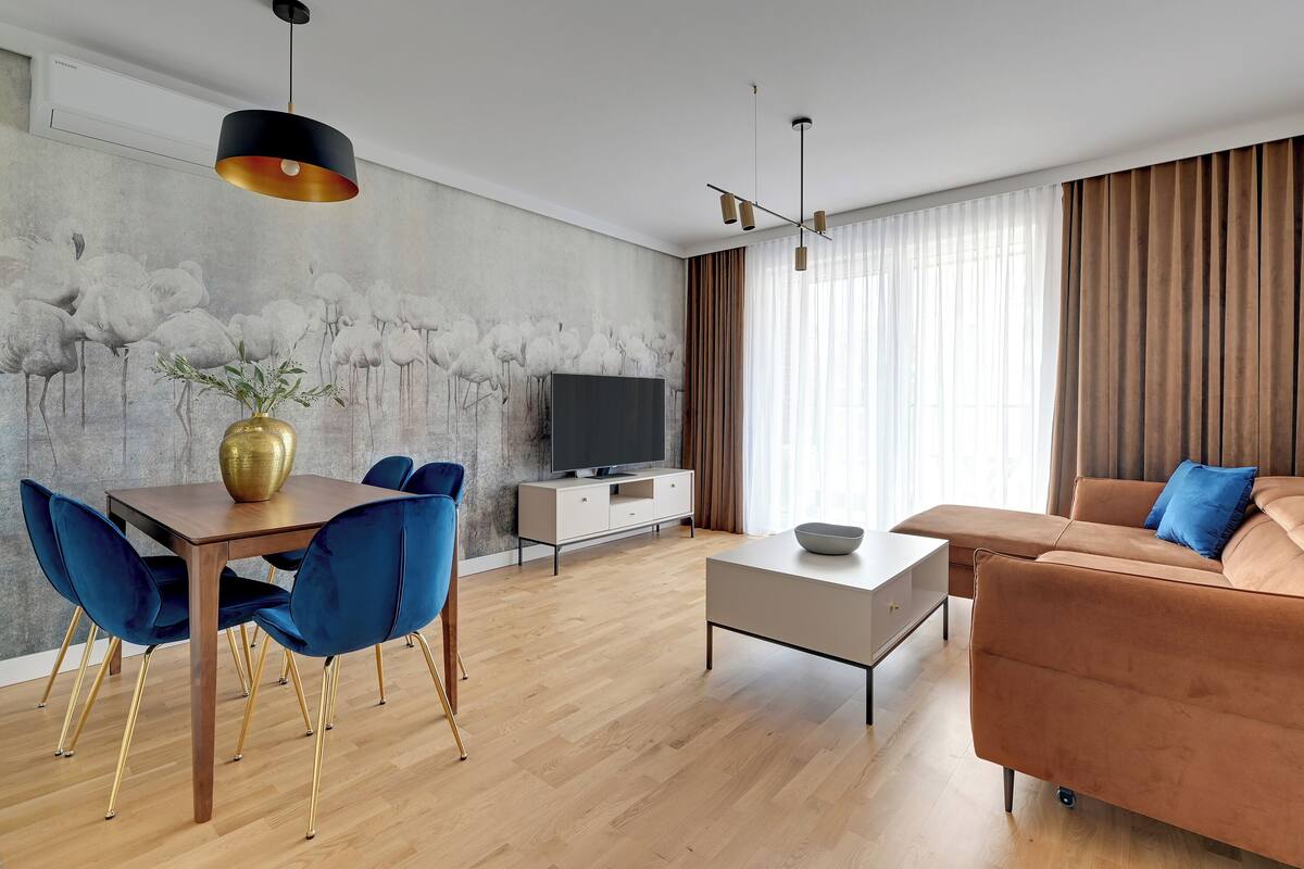 Uma sala de estar moderna com uma mesa de jantar de madeira, cadeiras azuis, um sofá e uma TV em um armário branco.
