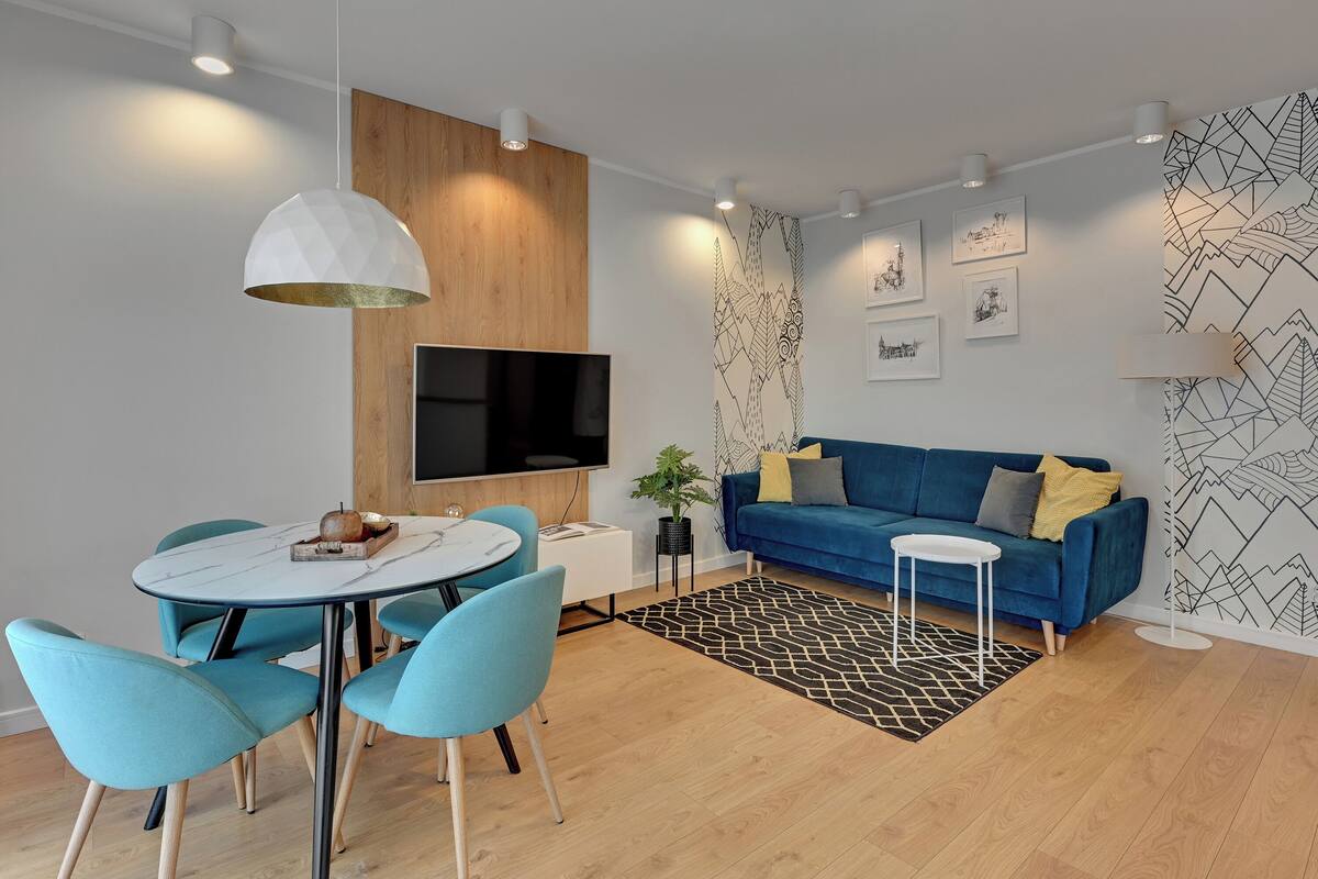 Uma sala de estar moderna com um sofá azul, uma mesa de jantar com cadeiras, uma TV fixada em um painel de madeira e arte decorativa na parede com padrão.