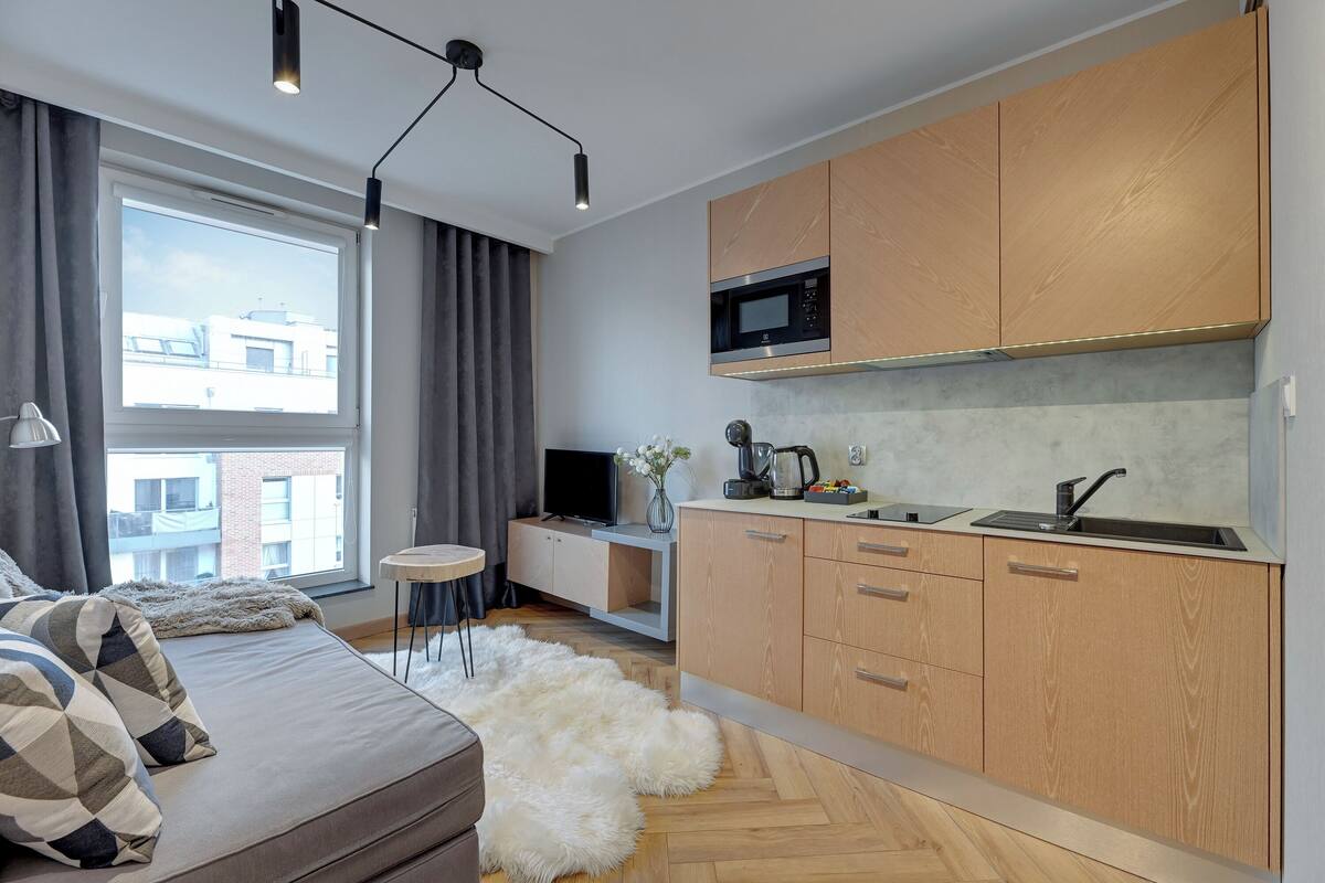 Um apartamento moderno com cozinha, um sofá com almofadas estampadas, um tapete felpudo branco e uma janela com vista para edifícios.