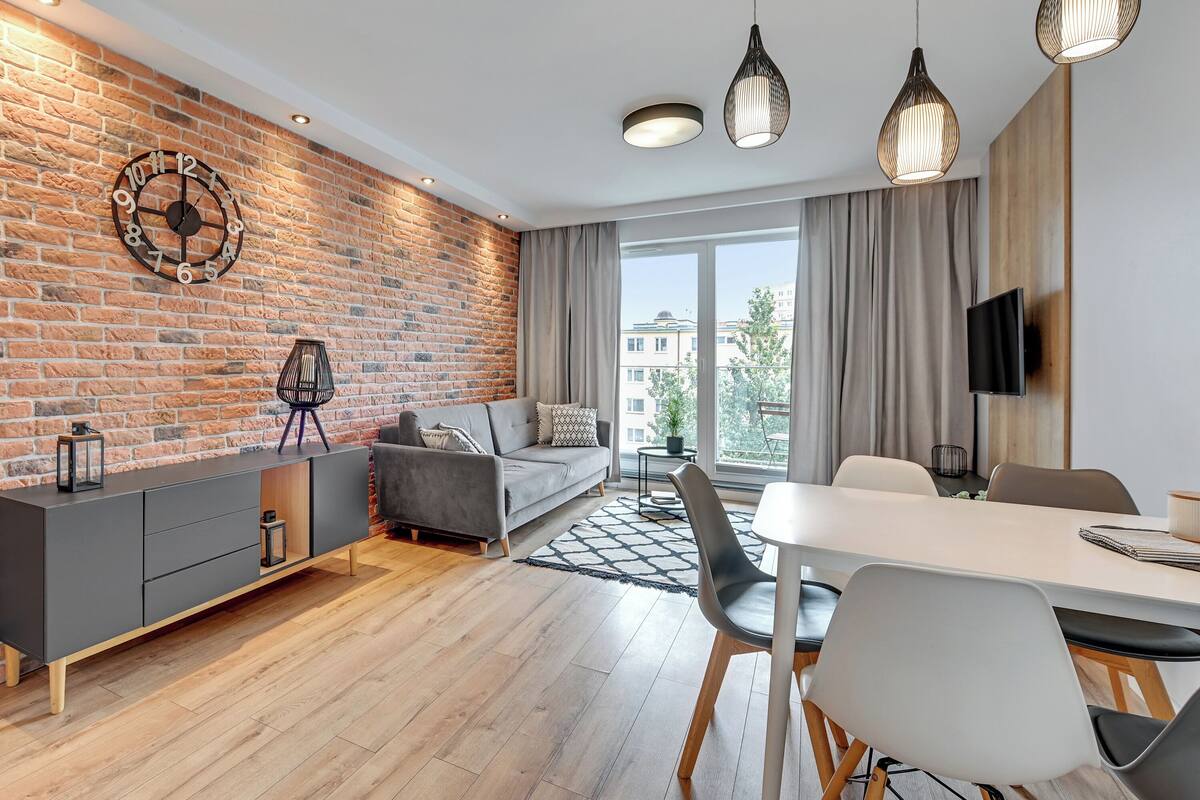 Uma sala de estar moderna com parede de tijolos, um sofá, uma mesa de jantar com cadeiras e uma TV fixada na parede.