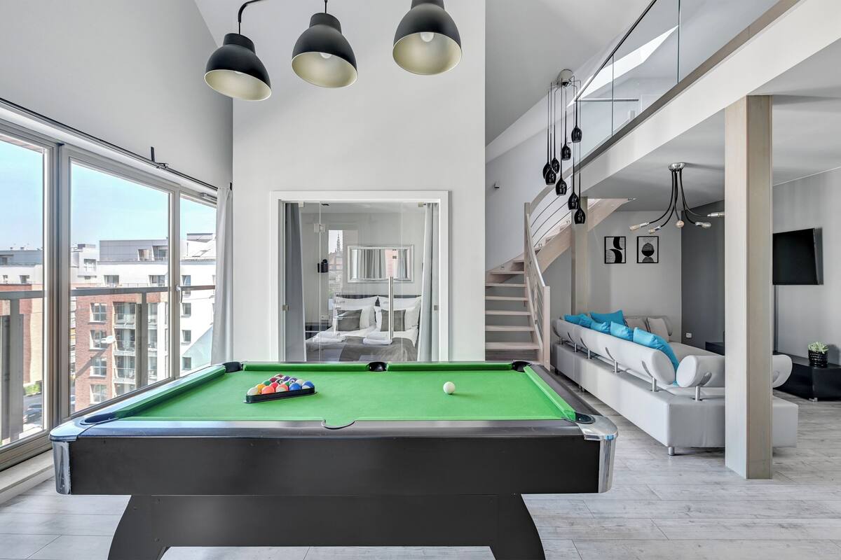 Uma sala de estar moderna com mesa de bilhar, janelas amplas, escada e sofá.