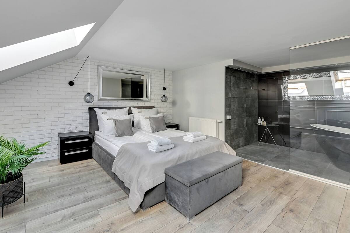 Quarto moderno com claraboia, uma cama grande com almofadas, mesa de cabeceira e uma pequena planta.