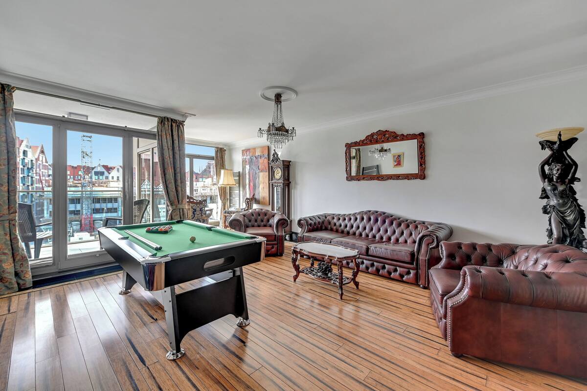 Uma sala de estar ampla com mesa de bilhar, sofás de couro e uma grande janela com vista para a cidade.
