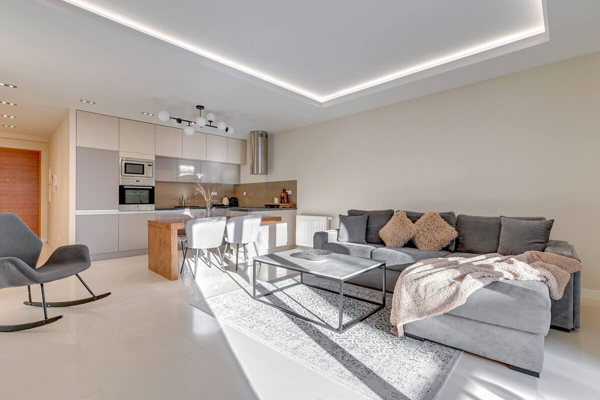 Uma sala de estar moderna com um sofá cinzento, uma mesa de centro e uma área de jantar com uma mesa e cadeiras brancas.