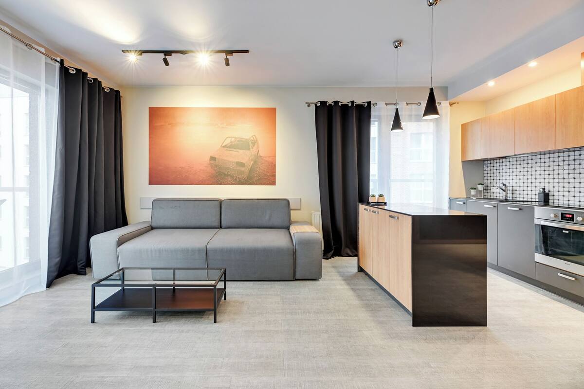 Um apartamento moderno com sofá cinzento, uma pequena mesa de centro, cozinha com armários de madeira e uma janela grande com cortinas.