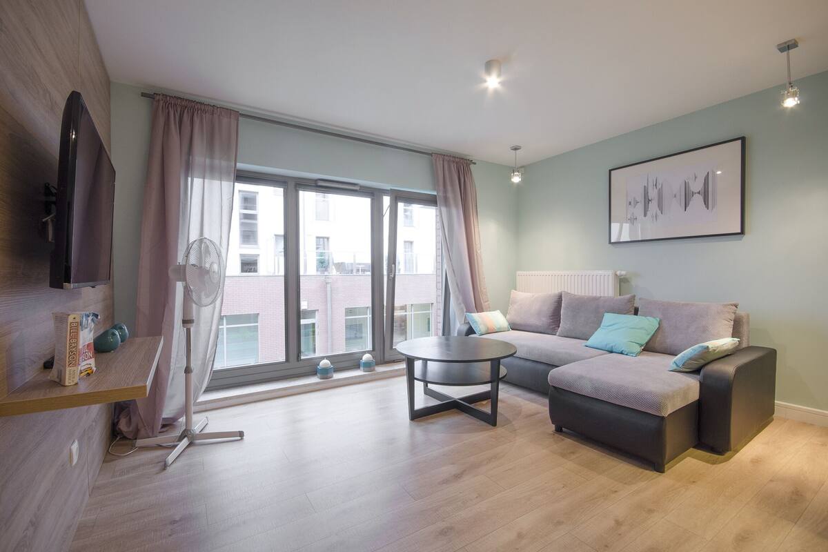 Uma sala de estar moderna com sofá, mesa de centro e televisão. Há uma janela com cortinas, um radiador e um quadro na parede.