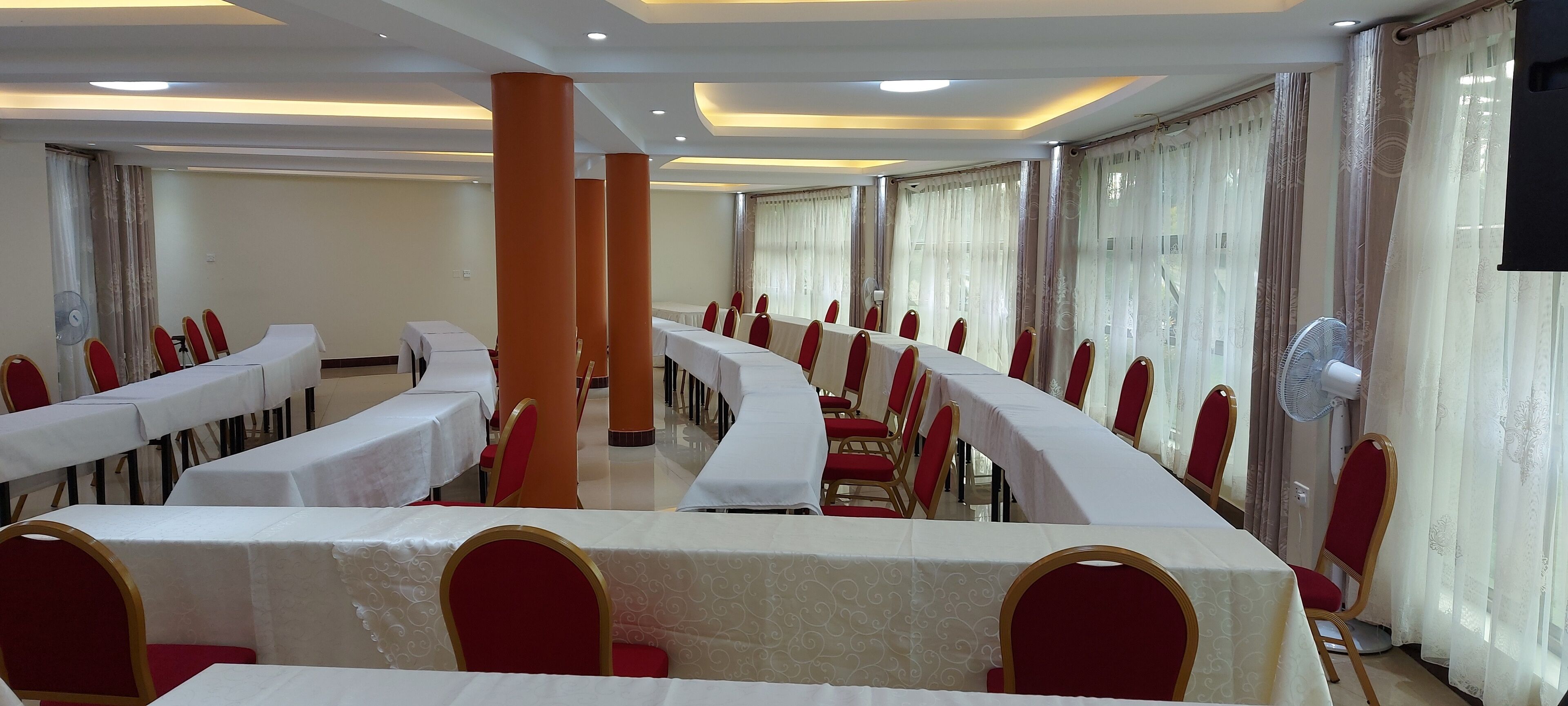Sala de conferências com várias mesas e cadeiras, pilares imponentes e um ventilador próximo às janelas.