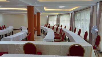 Sala de conferências com várias mesas e cadeiras, pilares imponentes e um ventilador próximo às janelas.