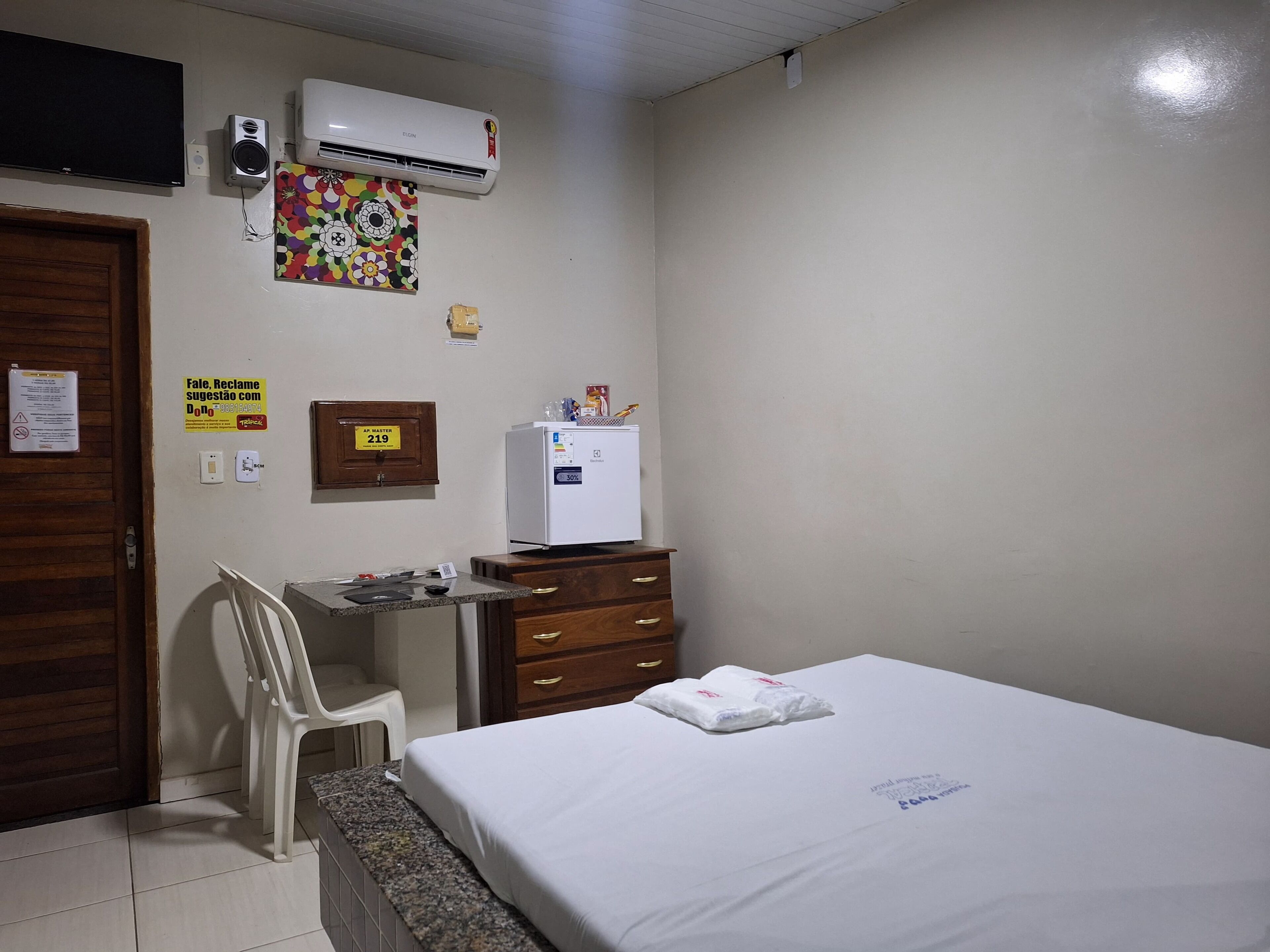 Quarto de hotel com cama, escrivaninha com cadeira, frigorífico, ar condicionado e televisão montada na parede.