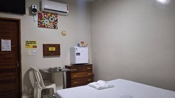 Quarto de hotel com cama, escrivaninha com cadeira, frigorífico, ar condicionado e televisão montada na parede.