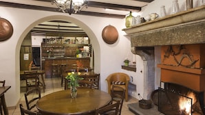 Uma área de refeições aconchegante com uma mesa de madeira redonda, cadeiras e um vaso com flores. Uma lareira com grade de metal é visível à direita.
