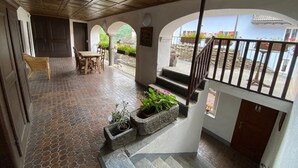 Uma área externa coberta com piso de azulejos, uma mesa de madeira com cadeiras, uma escadaria e plantas em vasos.