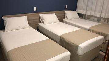 Quarto de hotel com três camas de solteiro, cada uma com colchão branco e manta bege. O quarto tem parede azul e cortina branca na janela.