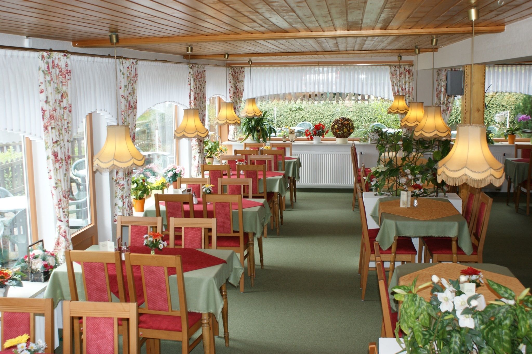 Um restaurante com mesas e cadeiras de madeira, cortinas florais e luminárias pendentes.