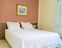 Cama bem arrumada com roupa de cama branca em quarto com paredes cor de pêssego e um quadro na parede.