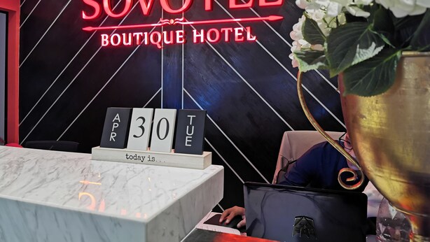 Bancada de recepção no Hotel Boutique SOVOTEL, com um letreiro de neon, um visor de data e um vaso grande de flores.