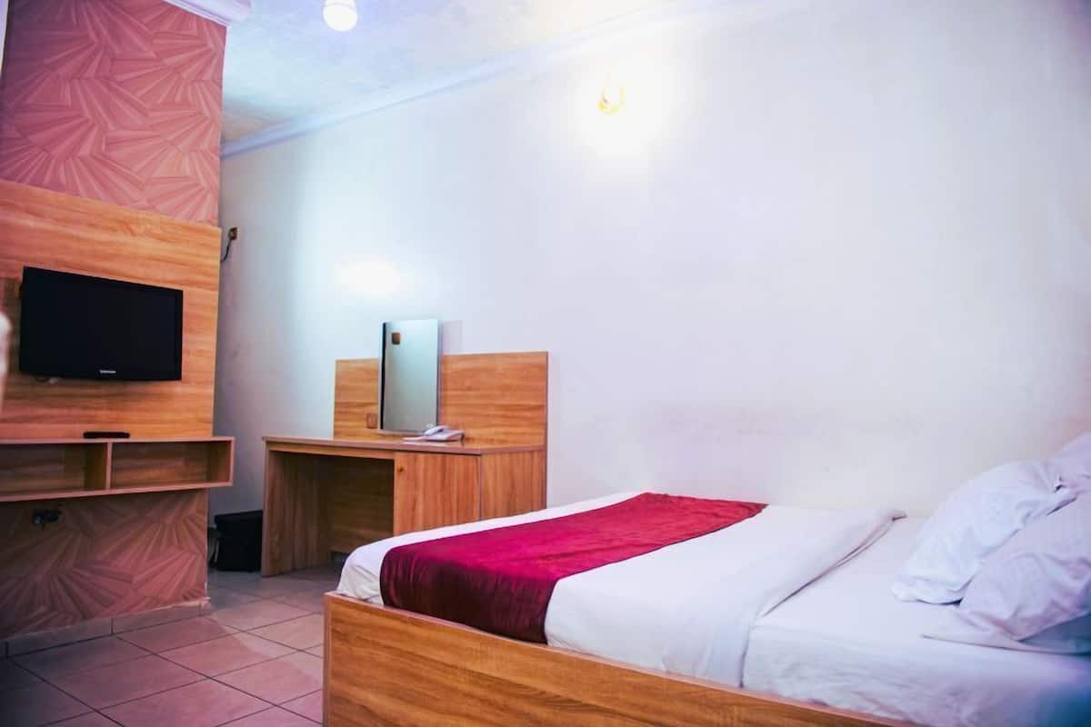 Quarto de hotel com cama de madeira, uma escrivaninha, televisão fixada na parede e um espelho.